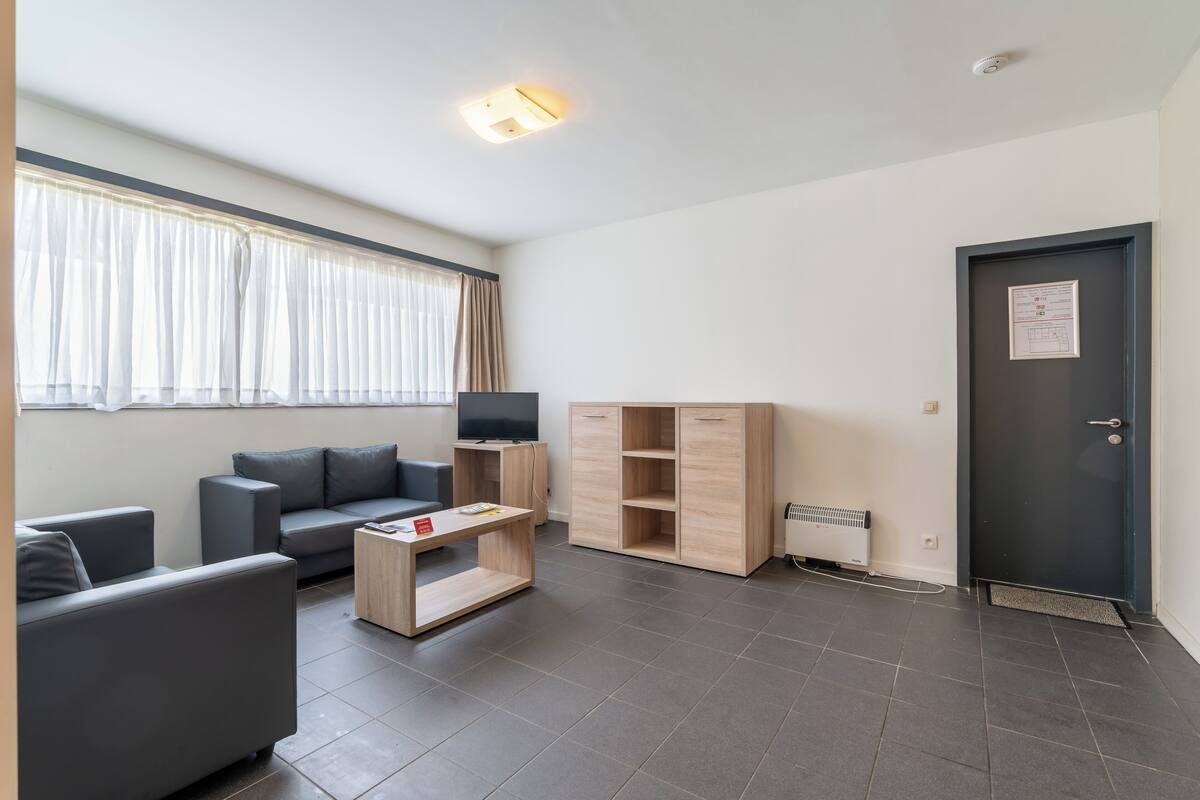 Uma sala de estar moderna com um sofá, uma mesa de centro e um suporte para TV. Há uma porta com um aviso e um radiador no chão.