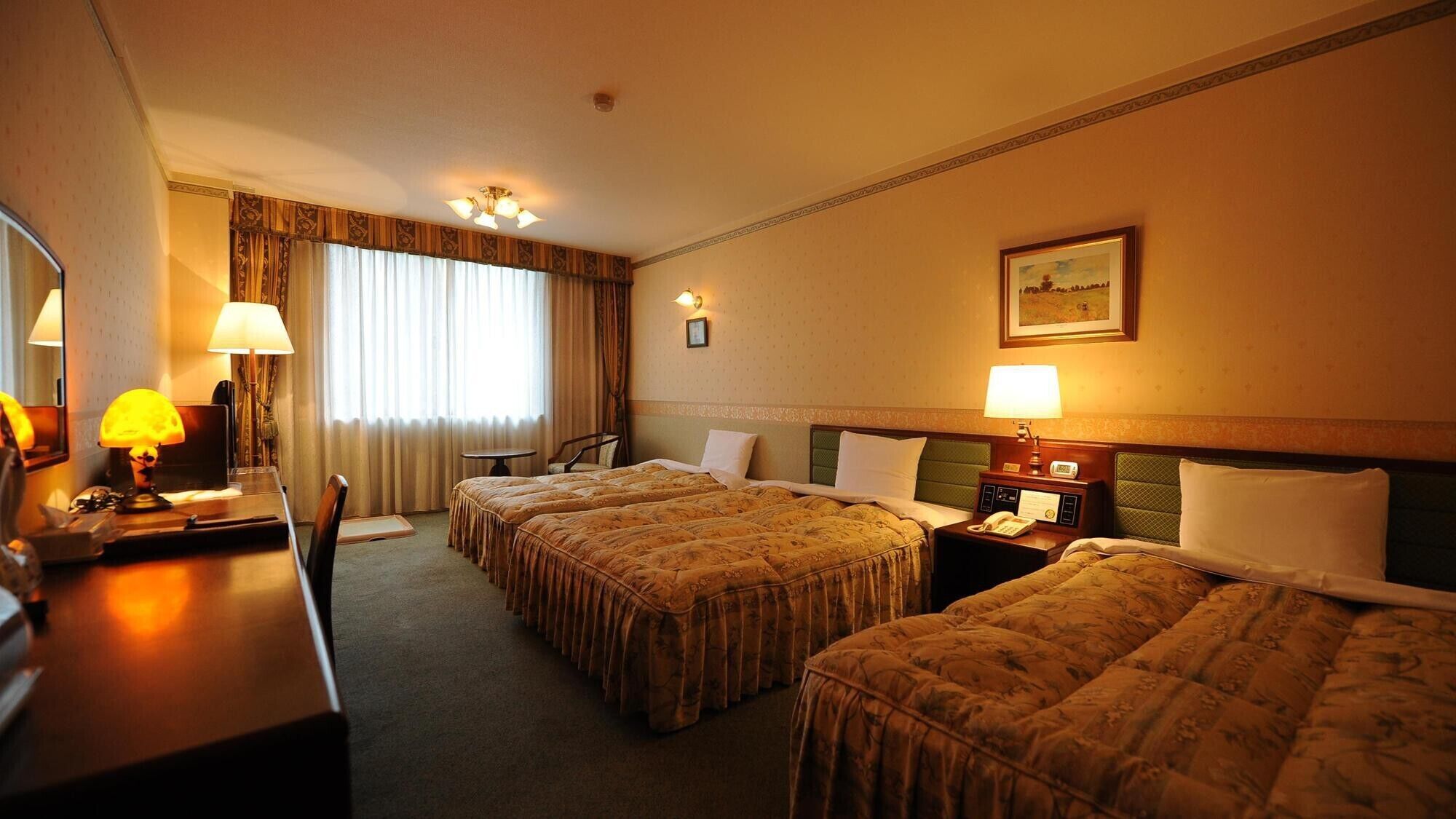 Quarto de hotel com duas camas, uma escrivaninha, uma cadeira, um abajur, uma mesinha de cabeceira e um quadro na parede.