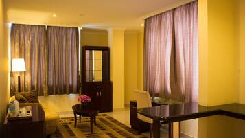 Quarto de hotel com sofá, mesa de jantar, mesa de centro e uma luminária. Há um espelho na parede e uma porta que dá para outro cômodo.