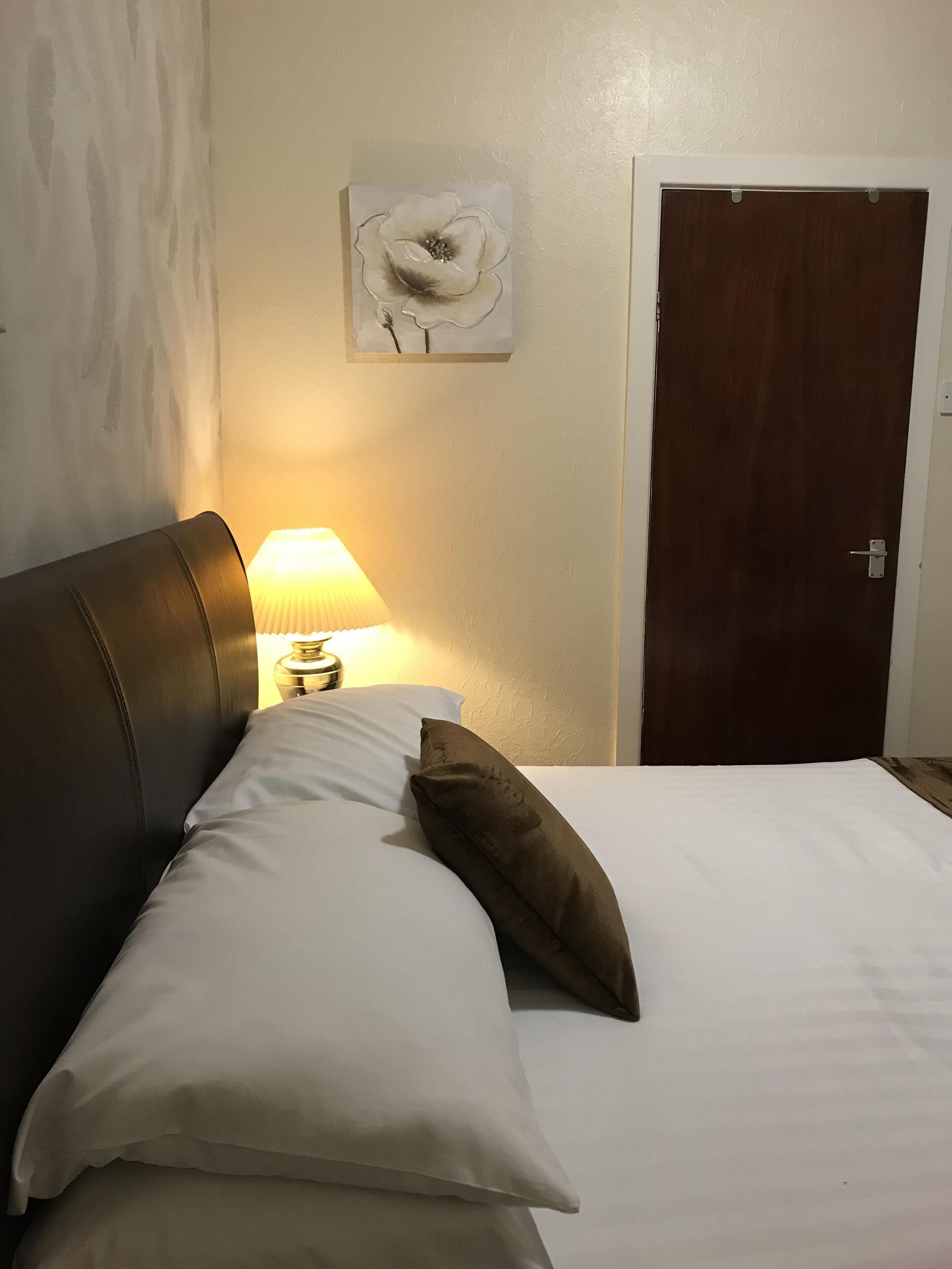 Quarto de hotel com cama, mesa de cabeceira com abajur, porta de madeira e uma obra de arte na parede.