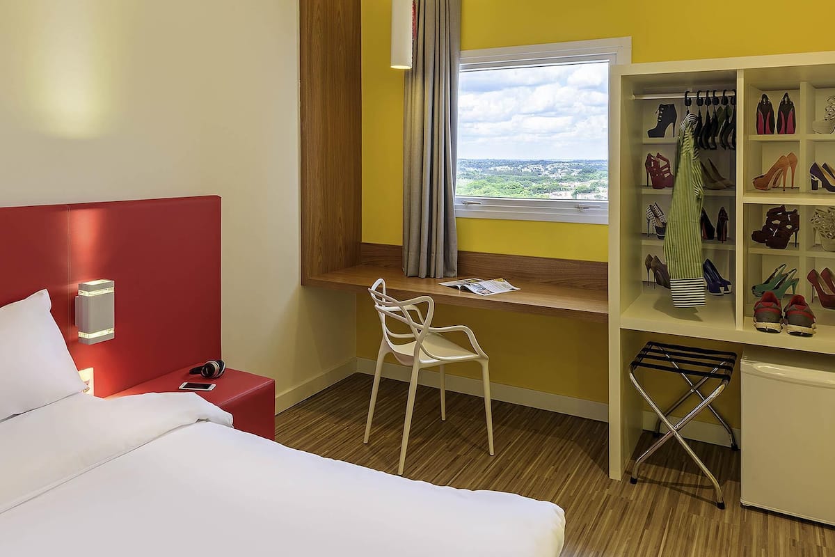 Quarto de hotel com cama, escrivaninha com cadeira, guarda-roupa com sapatos e janela com vista.