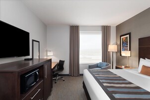 Quarto de hotel com cama, escrivaninha com cadeira, televisão, luminária e janela com cortinas.