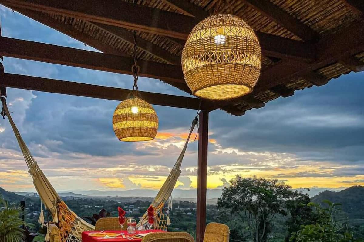 Uma área de estar ao ar livre acolhedora, com móveis de vime, uma mesa com toalha vermelha e luminárias de vime suspensas.