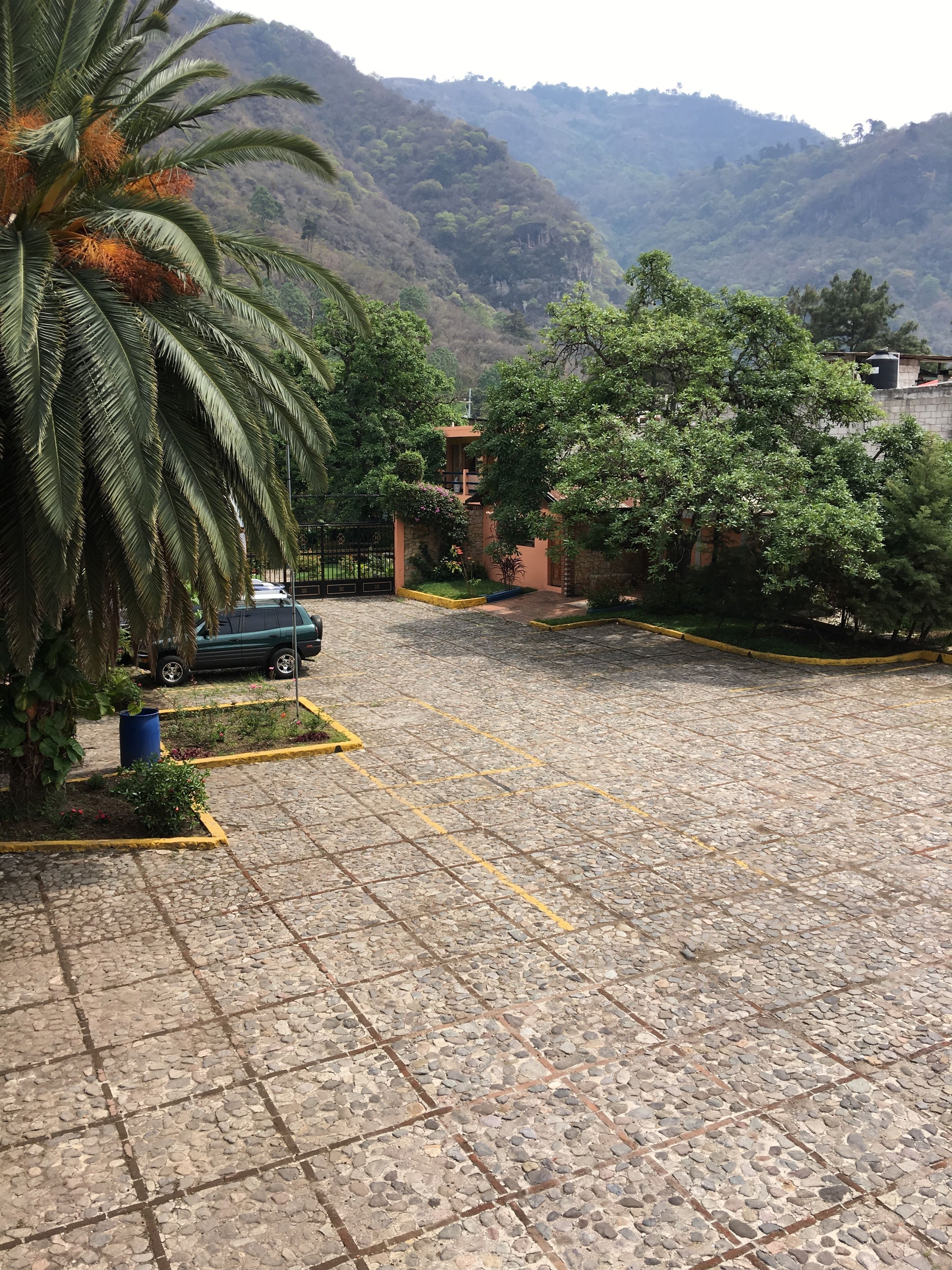 Pátio pavimentado com uma palmeira, um carro e um edifício com parede de pedra e um portão.