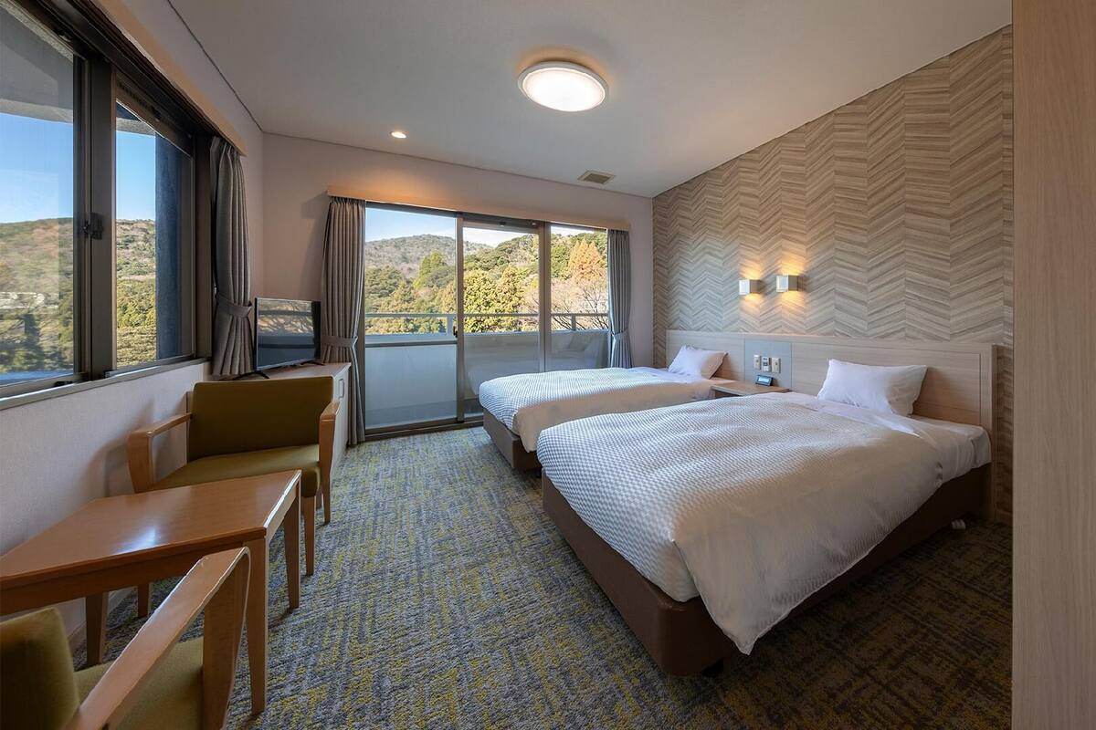 Quarto de hotel com duas camas, uma escrivaninha, uma cadeira, televisão e vista para o exterior.
