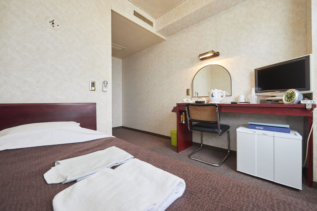 Quarto de hotel com cama, escrivaninha com computador, televisão e um pequeno frigorífico.