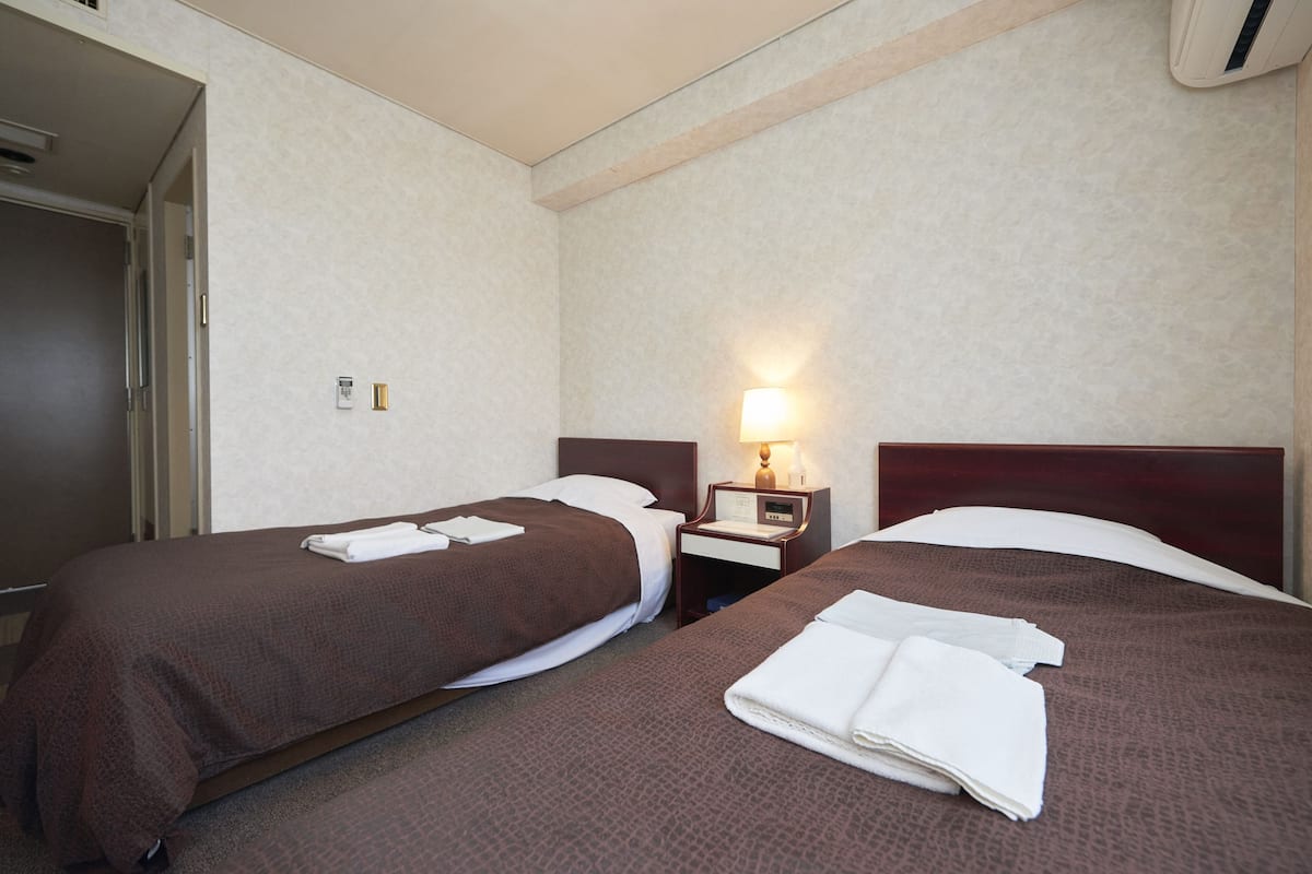 Quarto de hotel com duas camas, cada uma com colcha marrom e almofadas brancas. Há uma mesinha de cabeceira com uma luminária e uma pequena mesa entre as camas.