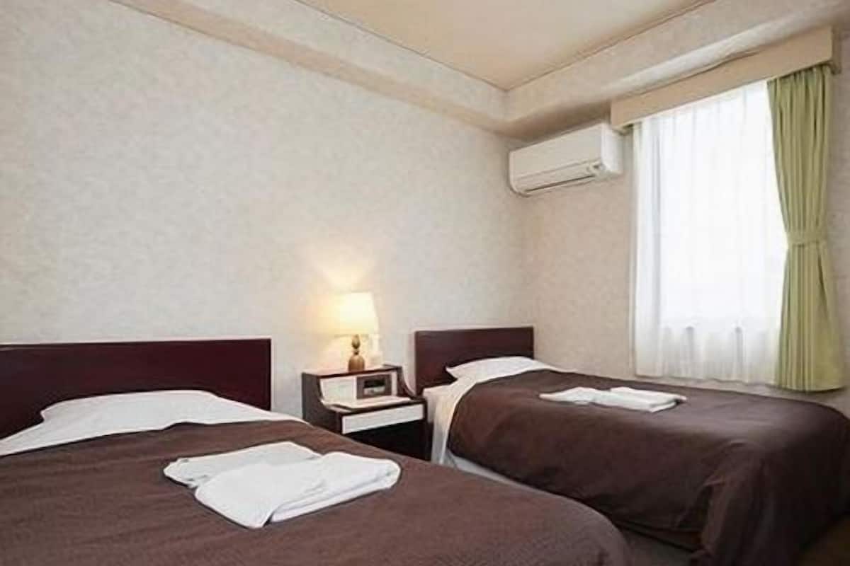 Quarto de hotel com duas camas, mesa de cabeceira com abajur, ar condicionado e janela com cortinas.