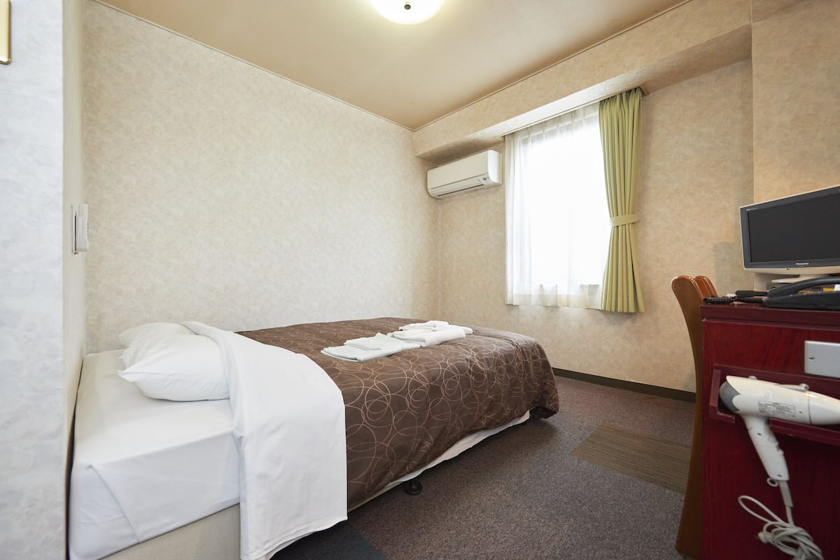 Quarto de hotel com cama, escrivaninha com computador, ar condicionado e janela com cortinas.
