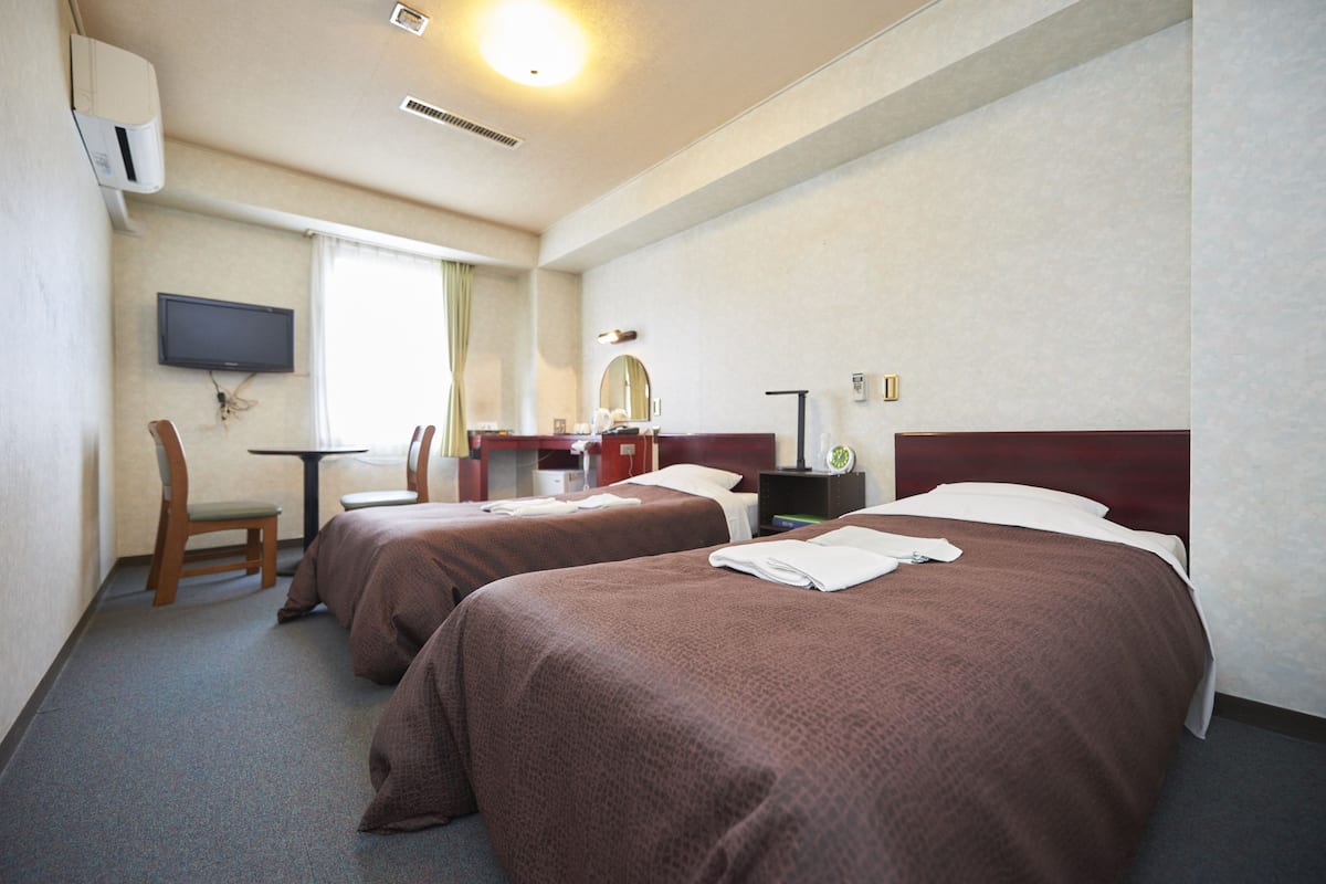 Quarto de hotel com duas camas, uma televisão, uma escrivaninha com cadeira e uma mesinha.