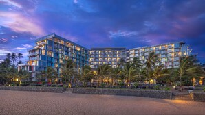 Hotel com vários andares e varandas, situado perto de uma praia com palmeiras.