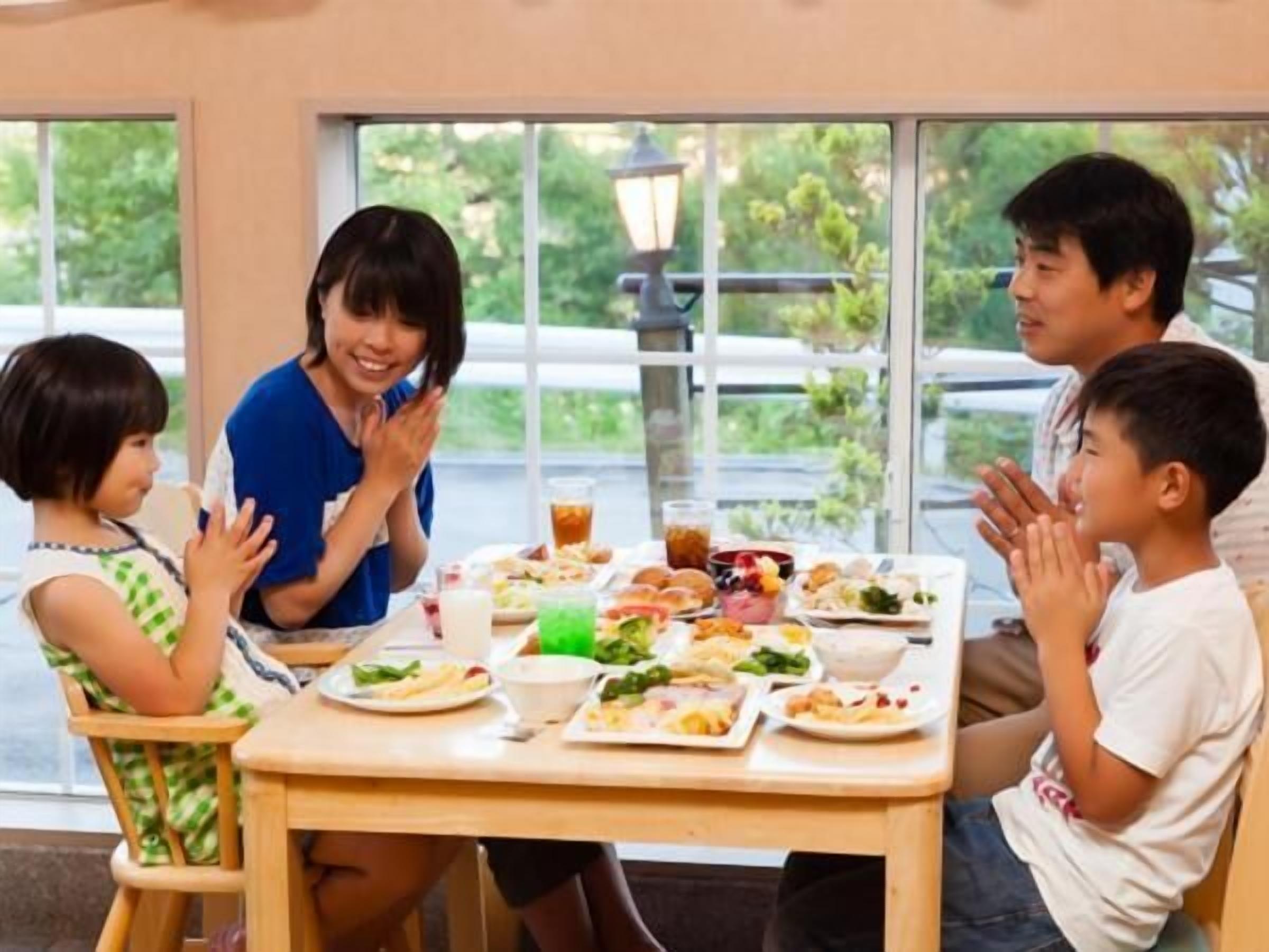 Uma família jantando à mesa com diversos pratos e bebidas, em um ambiente com grandes janelas e vista para o verde lá fora.