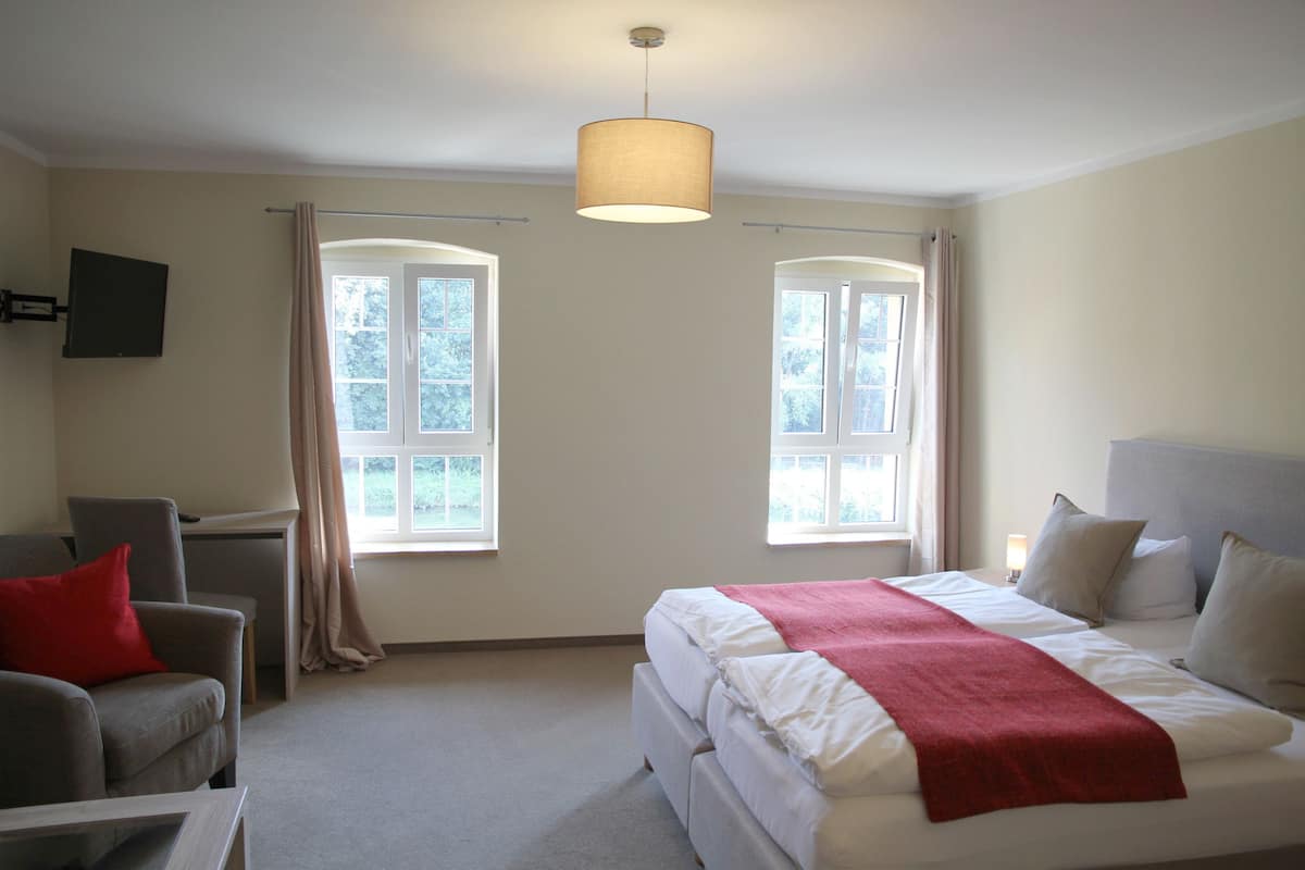 Quarto de hotel com cama, escrivaninha, cadeira e duas janelas.