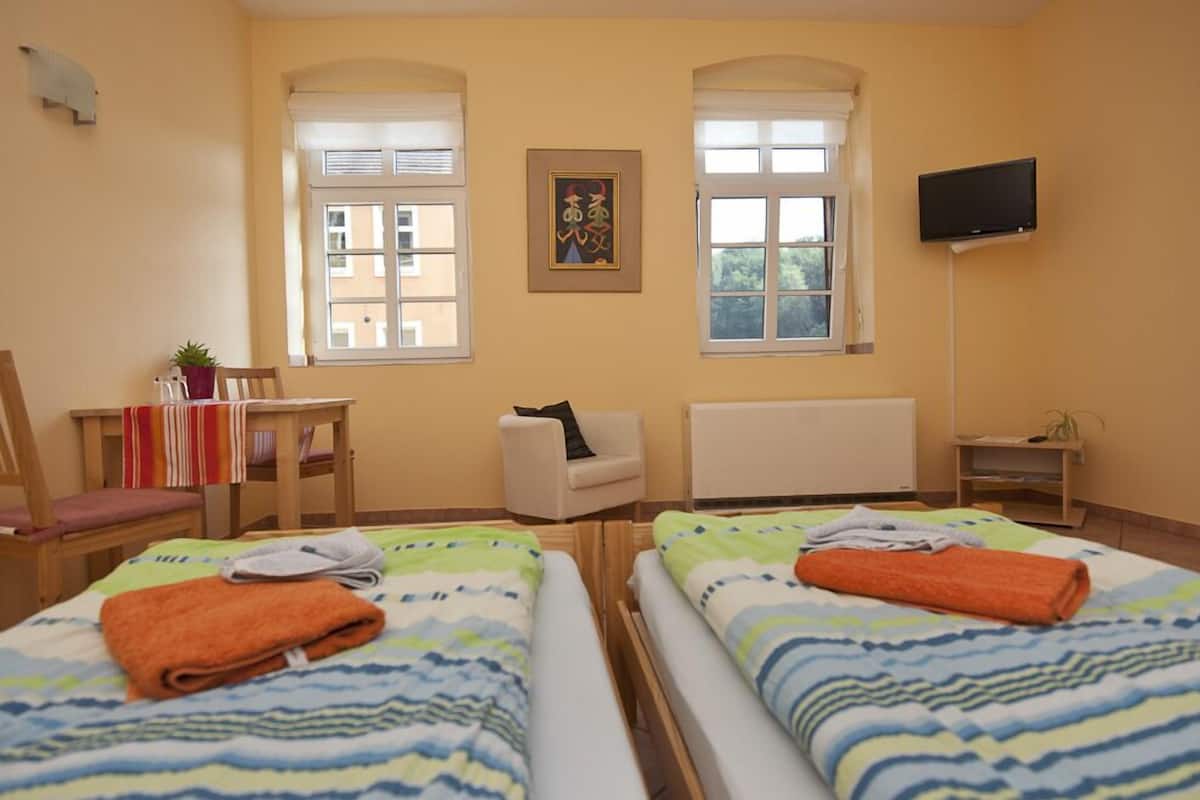Quarto de hotel com duas camas, uma cadeira, uma mesa pequena, televisão e janela.