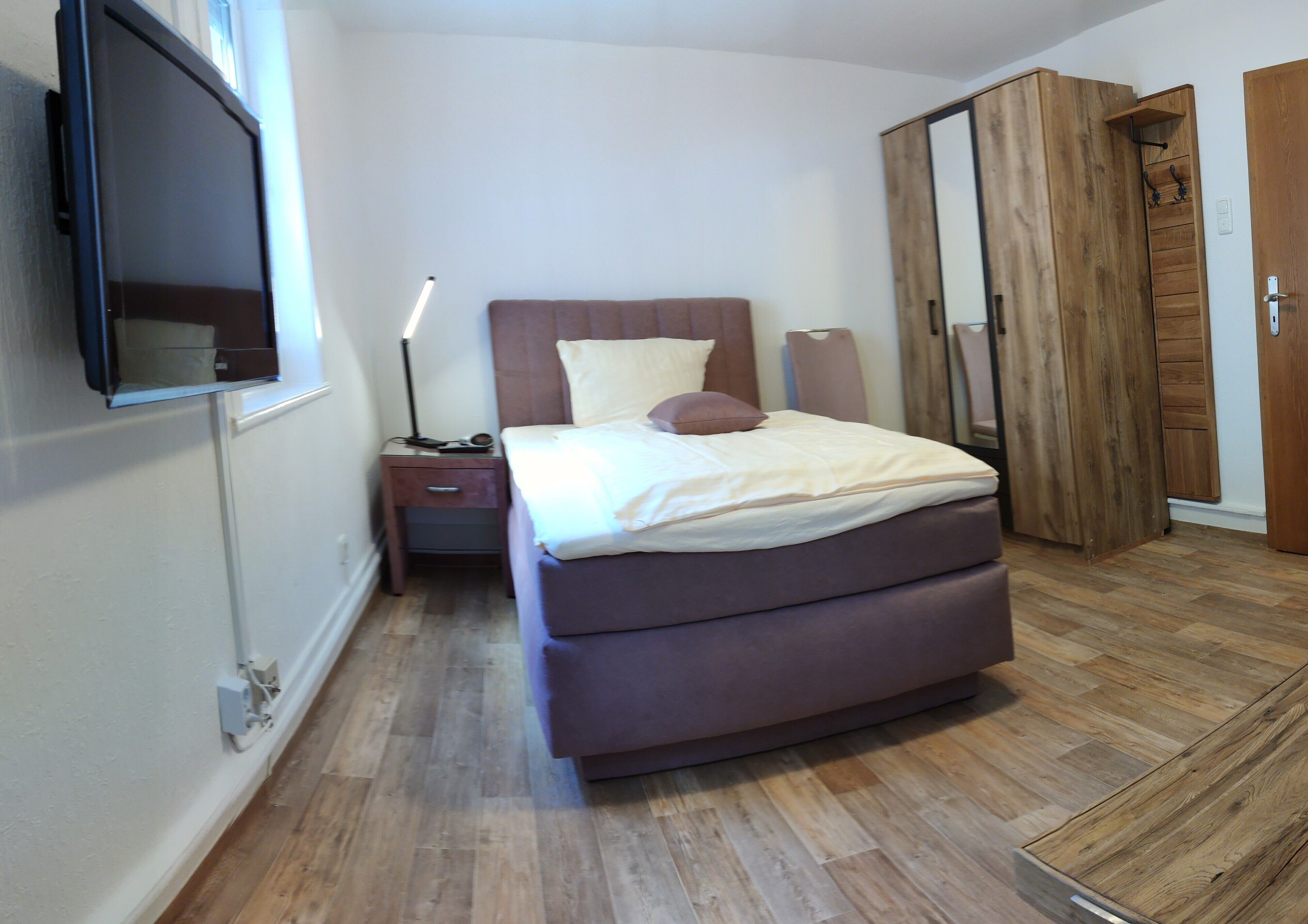 Quarto com cama, mesa de cabeceira, luminária, televisão, guarda-roupa e piso de madeira.