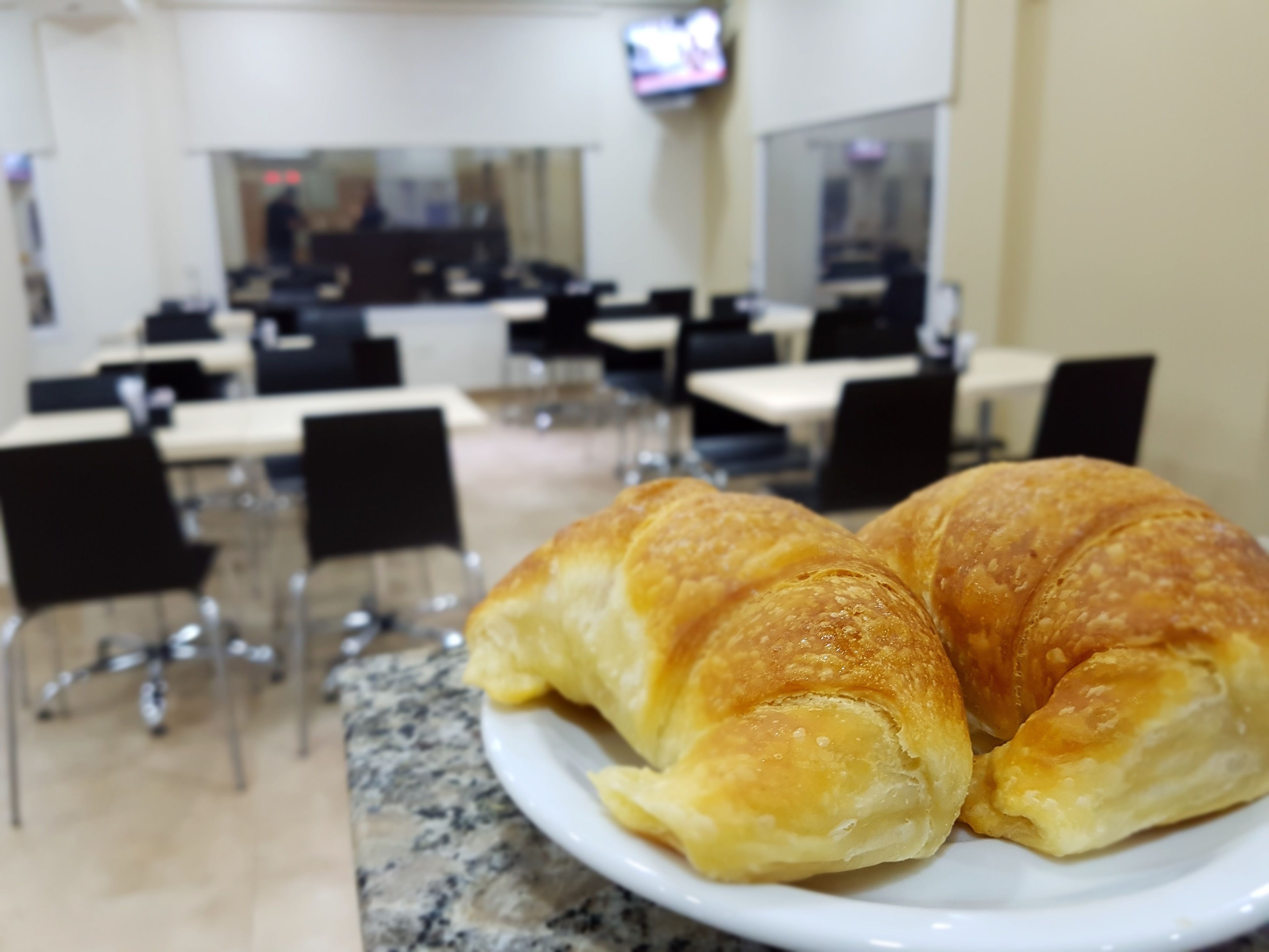 Uma área de refeições com mesas e cadeiras, uma televisão na parede e um prato de croissants em primeiro plano.