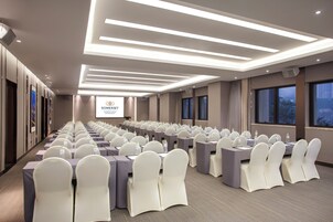 Sala de conferência com mesas e cadeiras dispostas em fileiras, uma tela de projetor e uma janela ampla.