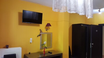 Quarto com parede amarela, televisão de tela plana montada acima de um espelho, pequena mesa com um vaso de flores, radiador e guarda-roupa preto.
