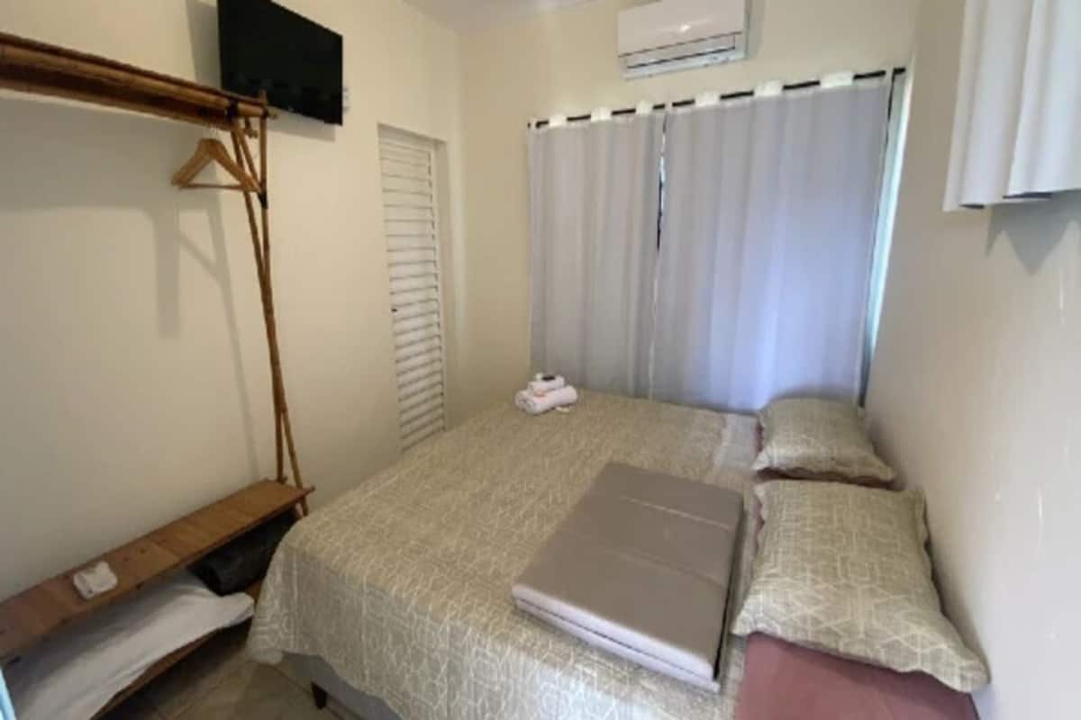 Quarto com cama, prateleira de madeira, televisão na parede e janela com cortinas.