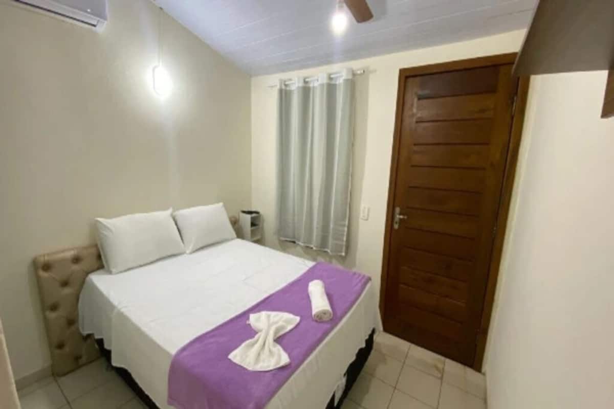 Quarto de hotel com uma cama, porta de madeira, janela com cortinas e um ventilador de teto.