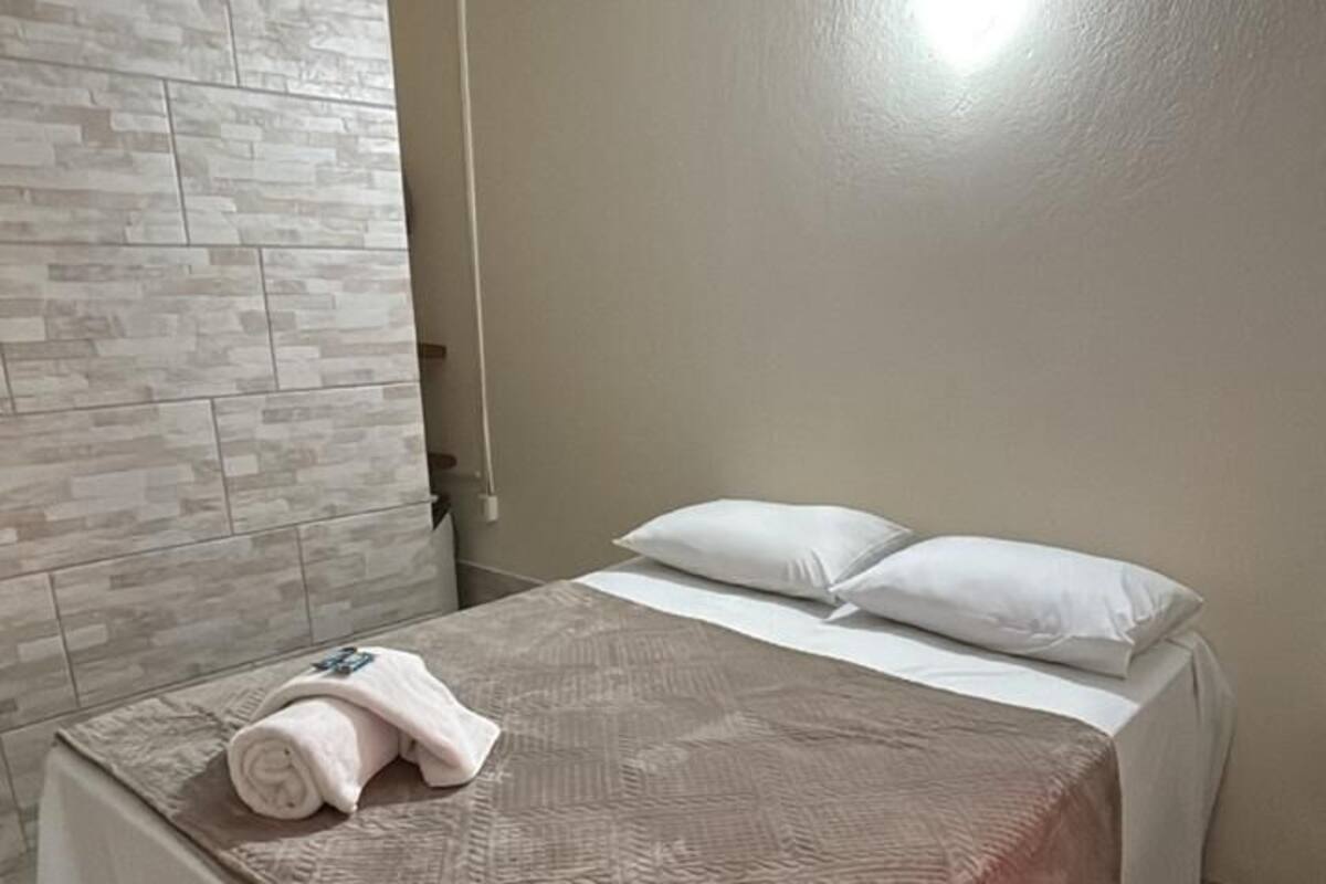 Uma cama de solteiro com colcha bege e marrom, almofadas brancas e uma toalha enrolada ao lado.