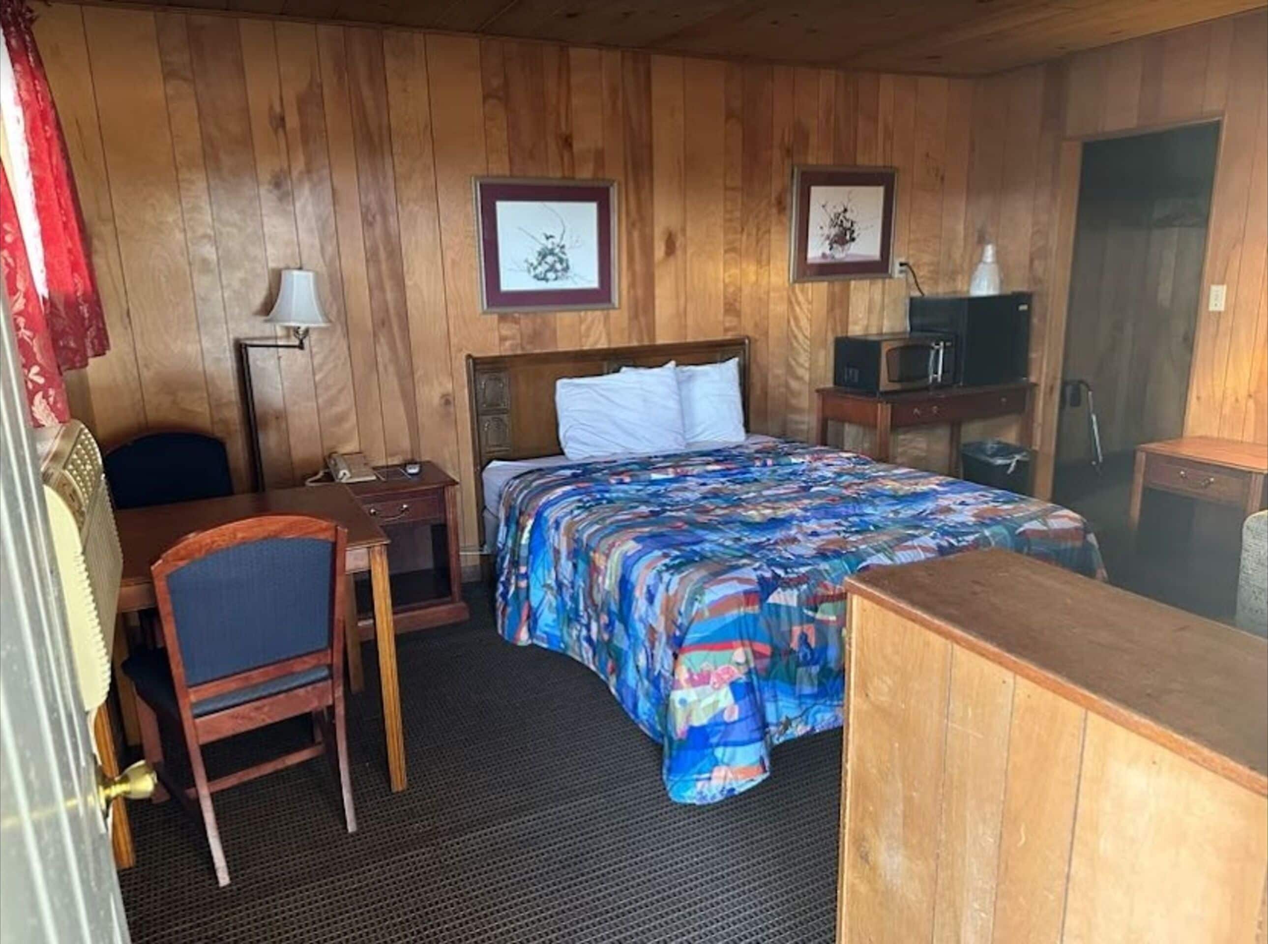 Quarto com painéis de madeira, uma cama, uma escrivaninha, uma cadeira, um abajur e uma televisão.