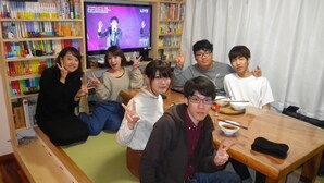 Um grupo de pessoas sentadas em uma sala com uma televisão exibindo um programa, cercadas por estantes de livros.