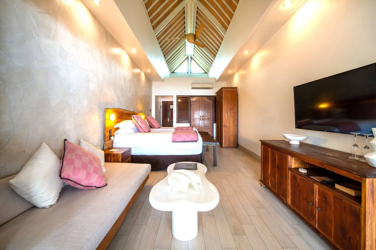 Quarto de hotel moderno com sofá, mesa de centro branca, armário de madeira, televisão e cama com cabeceira.