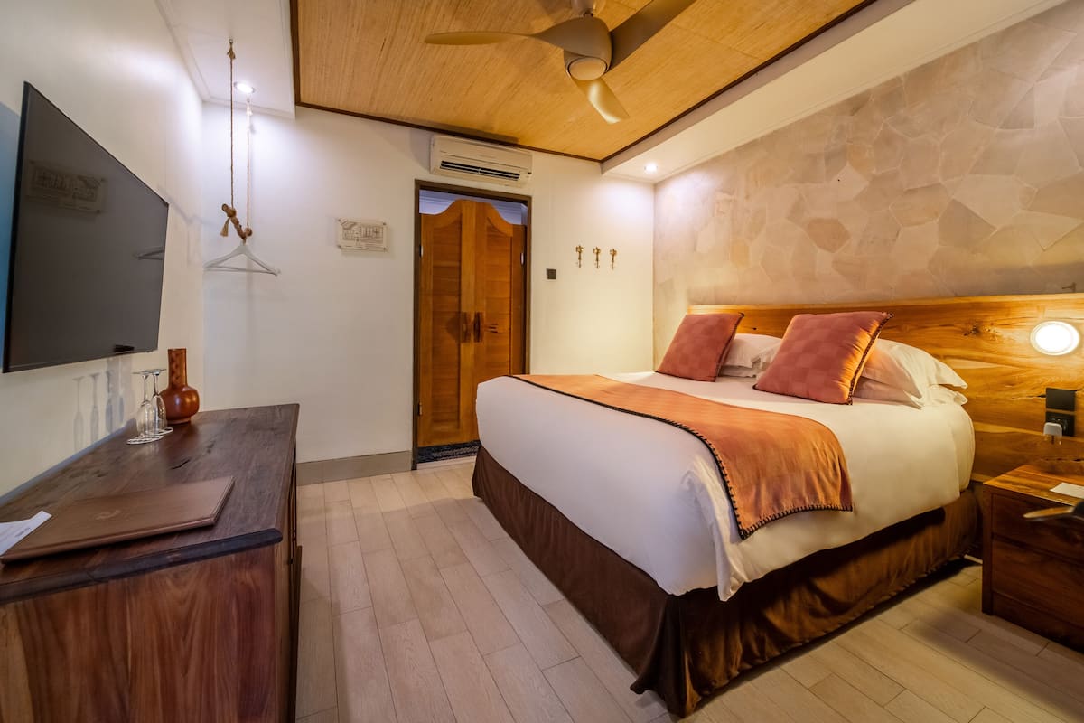 Quarto com cama, cabeceira de madeira, uma escrivaninha com um laptop, uma luminária pendente e uma parede de pedra.