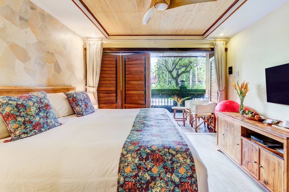 Quarto com cama grande, cabeceira de madeira, televisão e vista para o verde lá fora.