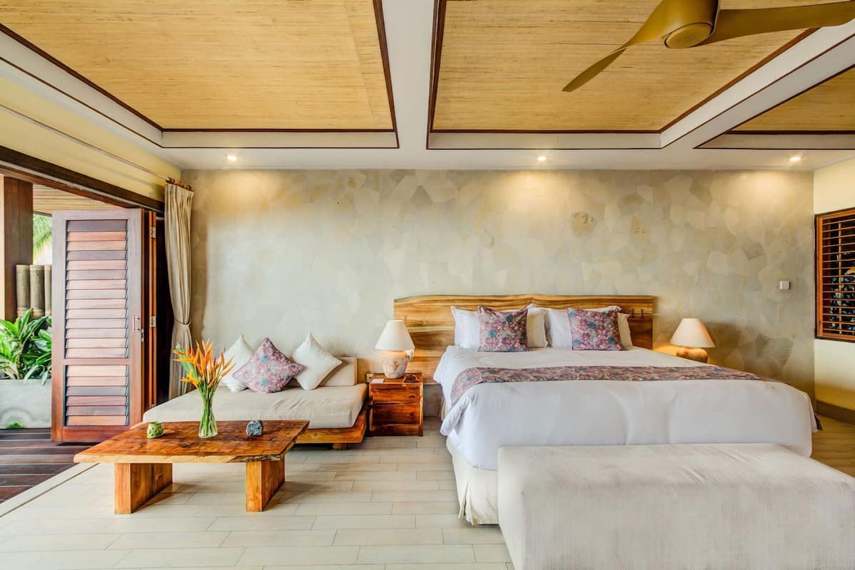 Quarto com cabeceira de madeira, cama com roupa de cama branca, mesa de cabeceira de madeira, mesinha de cabeceira com abajur e um banco de madeira.