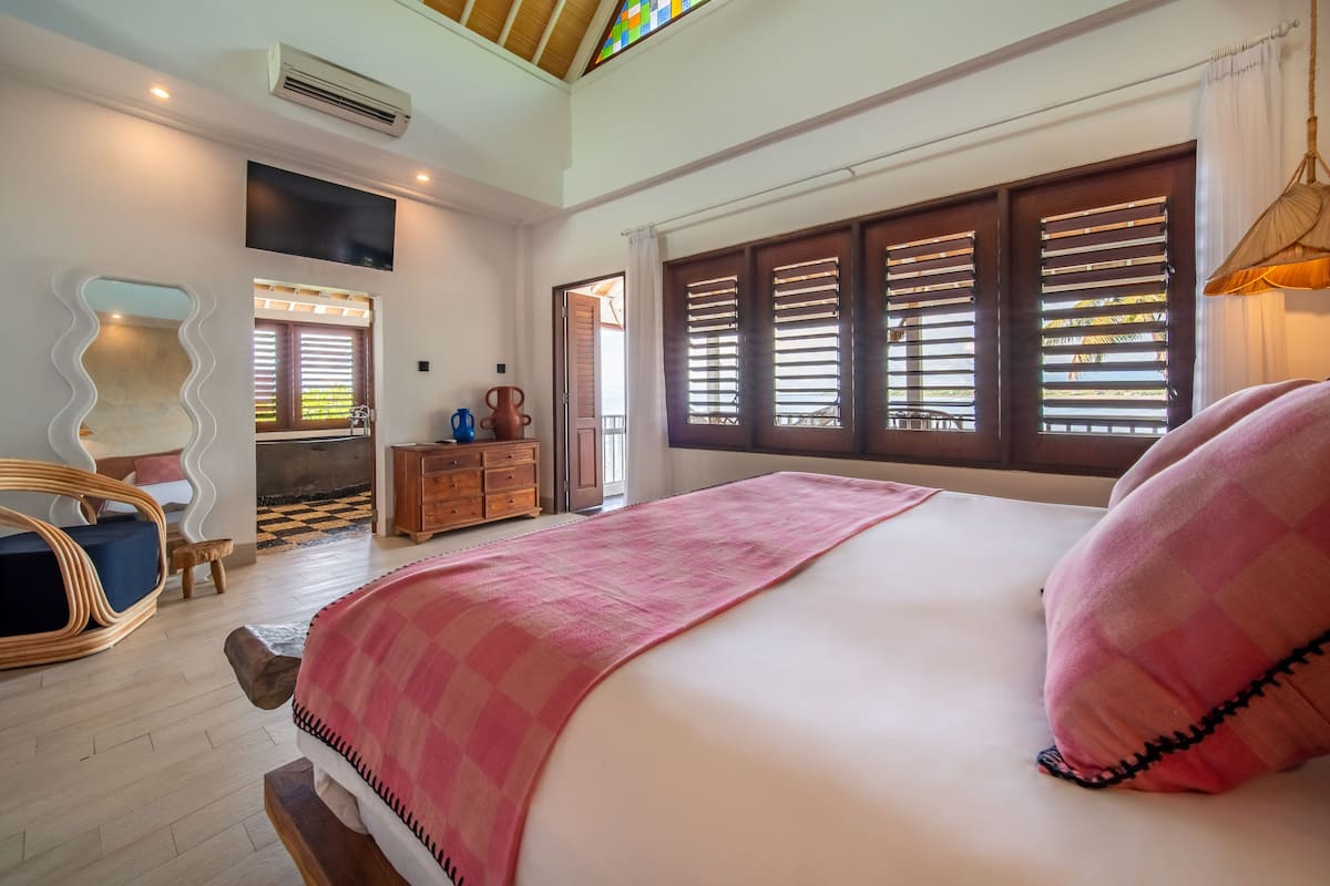 Quarto com uma cama grande, persianas de madeira, uma televisão e um espelho ondulado peculiar.