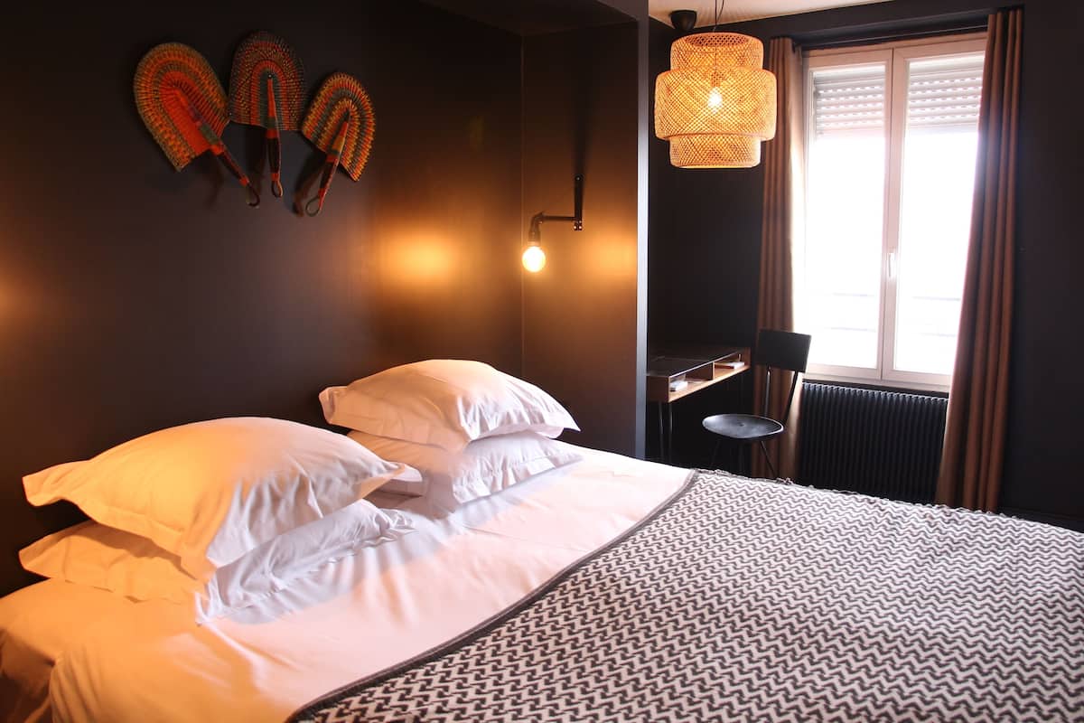 Quarto de hotel com cama, almofadas brancas, manta estampada, luminária na parede e janela com cortinas.
