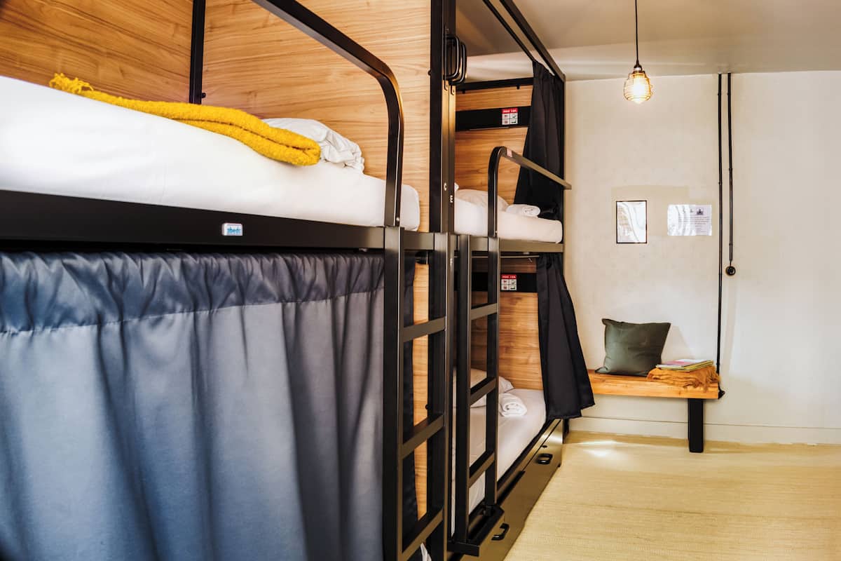 Quarto com beliche, cabeceira de madeira, um banco com almofada e luminária pendente.