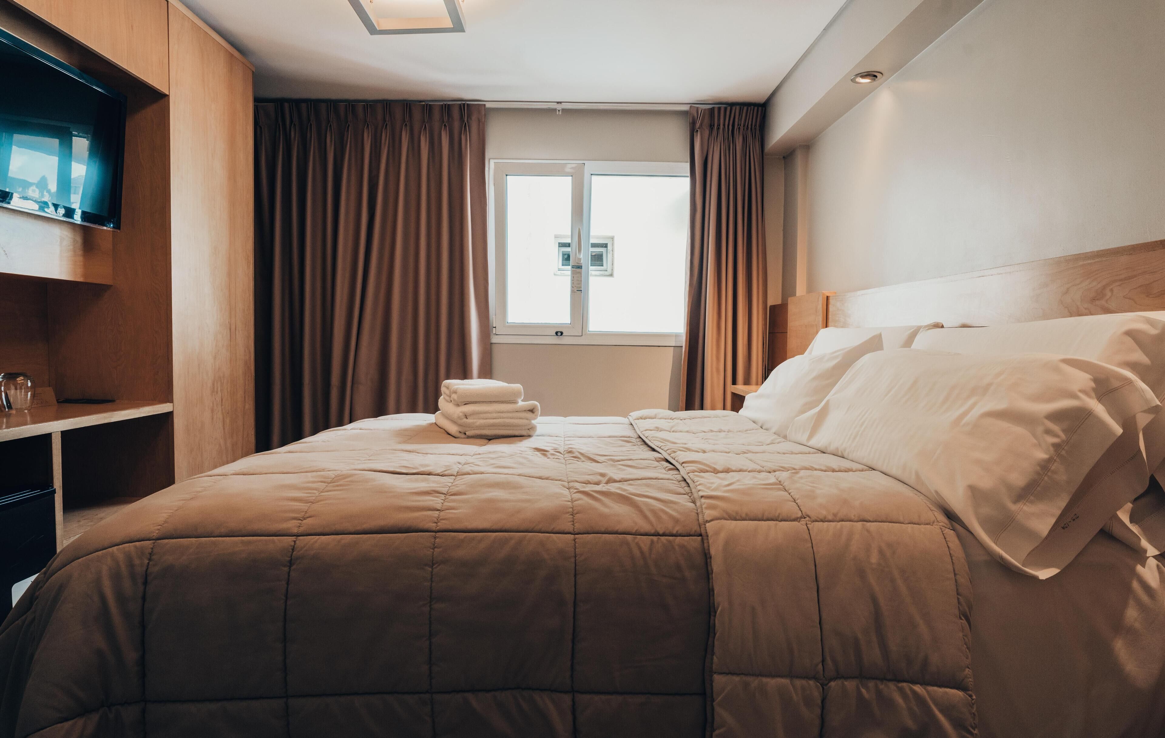 Quarto de hotel com uma cama grande, televisão fixada na parede e uma janela com cortinas.