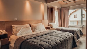 Quarto de hotel com duas camas, mesa de cabeceira com telefone e janela com cortinas.