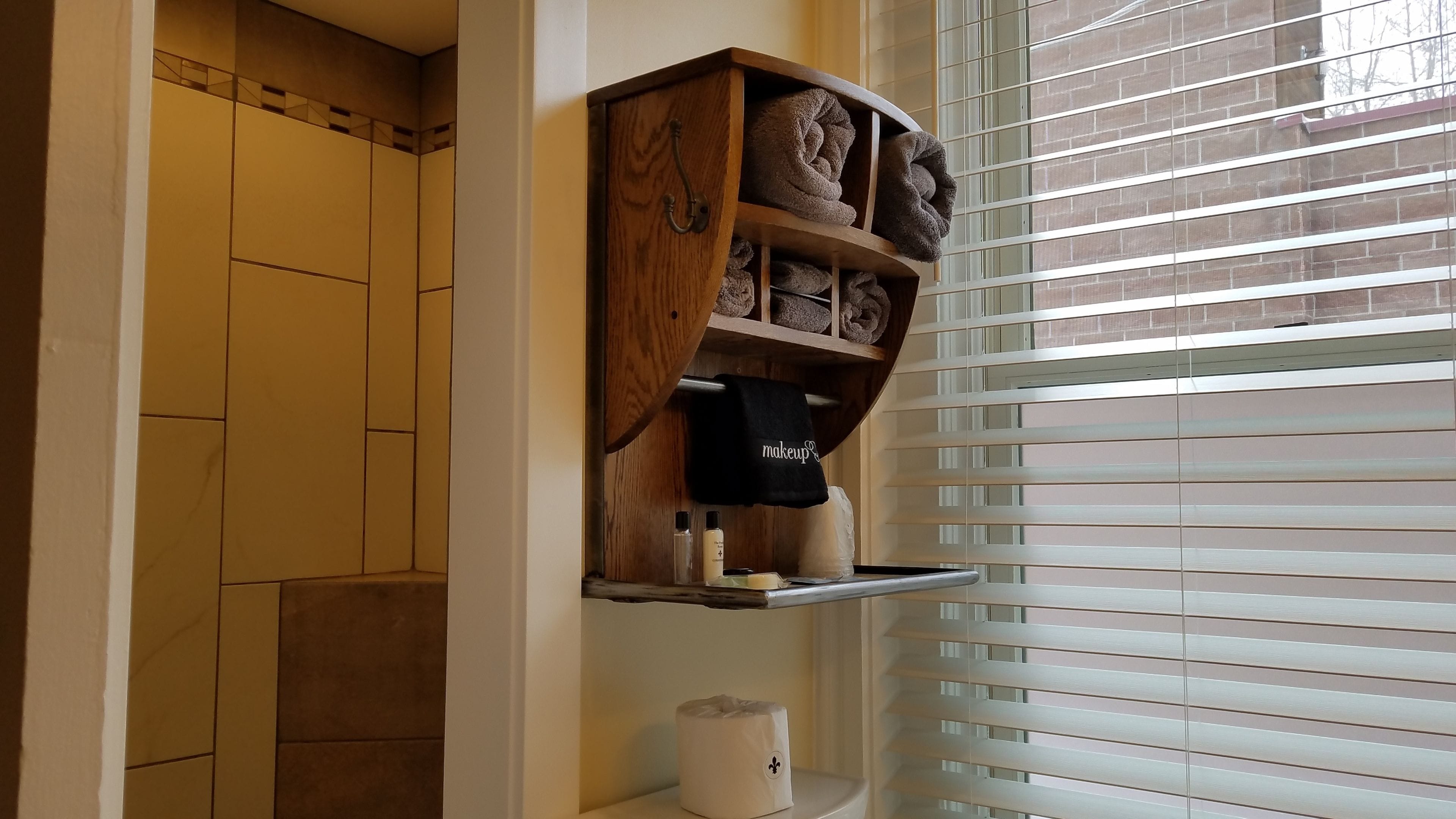 Casa de banho com uma prateleira de madeira com toalhas e produtos de higiene, um suporte de papel higiénico e uma janela com persianas.