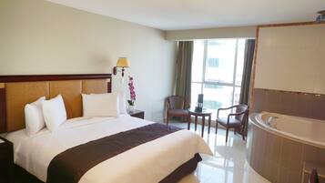 Quarto de hotel com uma cama grande, área de estar com uma poltrona e uma mesa pequena, banheiro com banheira e janela com cortinas.