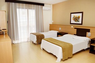 Quarto de hotel com duas camas, uma escrivaninha e uma janela grande com cortinas leves.