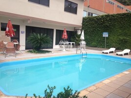 Uma piscina com espreguiçadeiras e guarda-sóis, cercada por um edifício com um pátio ladrilhado e um viveiro.