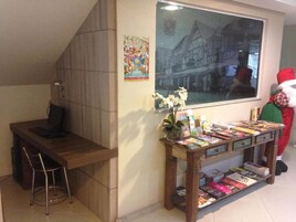 Uma área de recepção com uma mesa de madeira, uma cadeira, um vaso com flores e uma mesa expositora com livros e revistas.