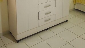 Um guarda-roupa branco com várias gavetas e portas, localizado num quarto com soalho em azulejos e uma mesa coberta por um pano amarelo.
