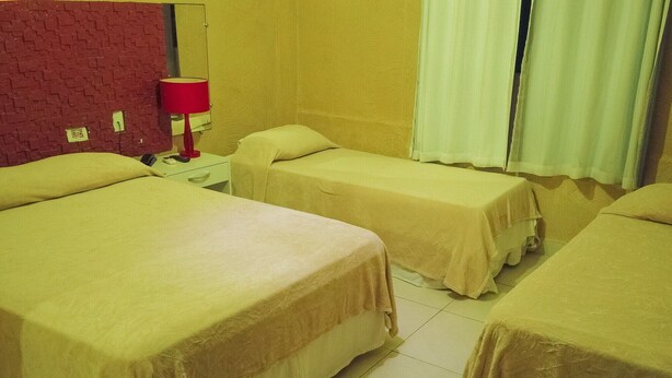 Quarto de hotel com quatro camas de solteiro, uma luminária vermelha, uma mesa pequena e um espelho na parede.