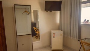 Quarto pequeno com guarda-roupa, televisão na parede, frigorífico e janela com cortinas.