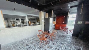Um espaço amplo e interno com piso de azulejos, mesas e cadeiras de madeira, e uma área de cozinha ao fundo.