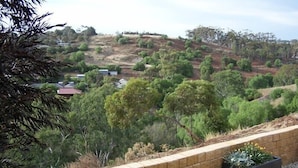 Uma área externa ajardinada com um muro de tijolos, plantas em vasos e vista para uma paisagem arborizada com colinas e edifícios ao longe.
