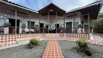 Um pátio com um caminho central de tijolos, rodeado por um edifício com várias portas e janelas, e plantas em vasos.
