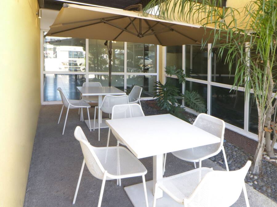 Um pátio externo com mesas e cadeiras brancas, um grande guarda-sol e um vaso de planta.