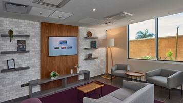Um hall de hotel moderno com parede de tijolos, painel de madeira, televisão de tela plana, sofás cinzas, mesa de centro e uma grande janela com vista para o exterior.