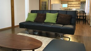 Sala de estar com um sofá azul escuro, almofadas verdes e marrons, uma mesa de centro de madeira e vista para a área da cozinha.